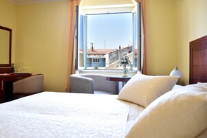Quarto de hotel com cama, escrivaninha, cadeira e janela com vista para prédios.