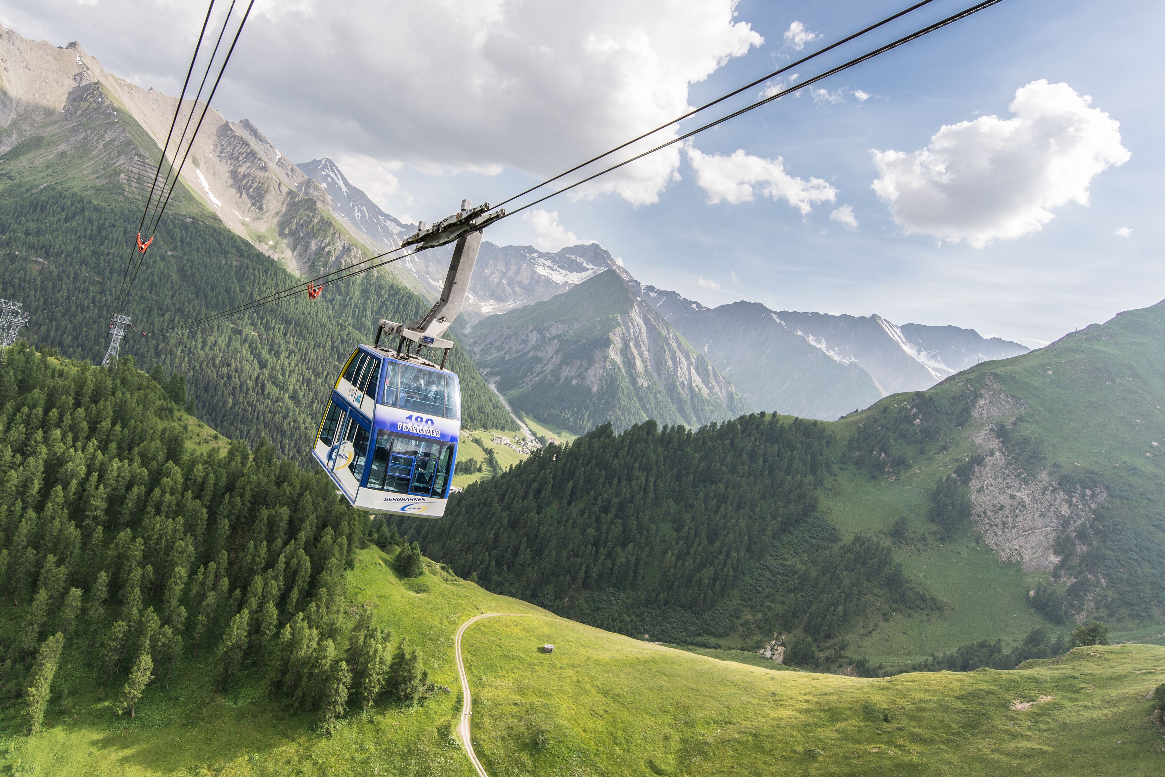 Teleférico a percorrer uma paisagem montanhosa com vales verdejantes e picos nevados ao longe.