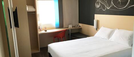 Quarto de hotel com cama, escrivaninha e cadeira. Há uma janela com vista para o exterior.