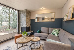 Uma sala de estar moderna com lareira de pedra, um espelho grande e vista para árvores através de uma janela ampla.