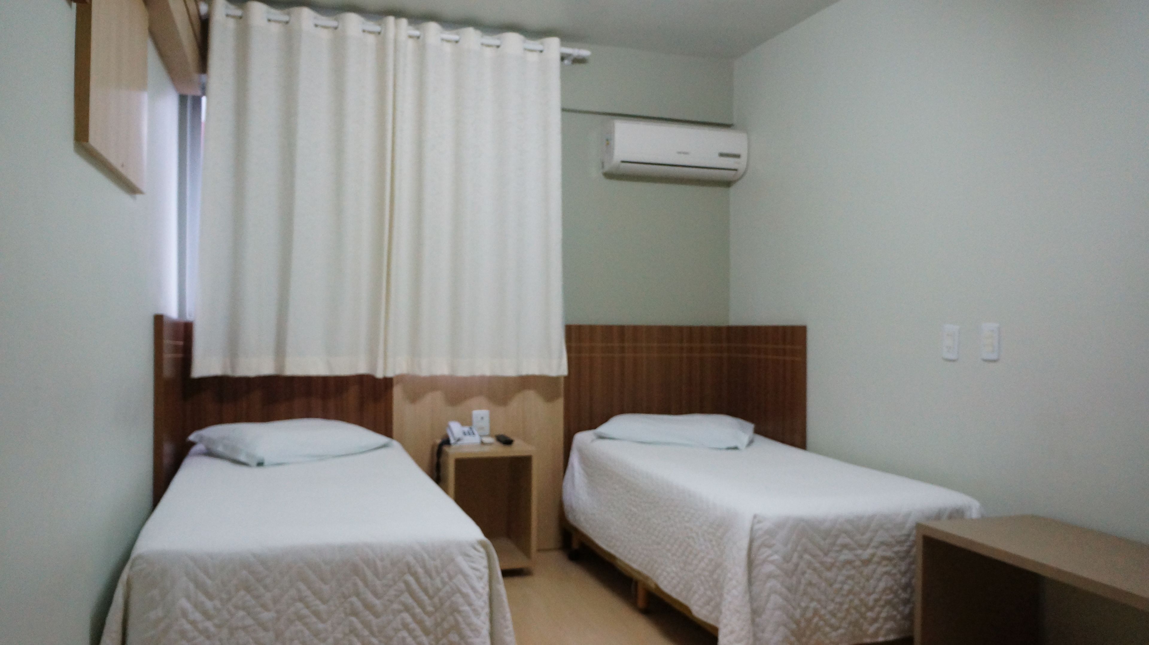 Quarto de hotel com duas camas de solteiro, cabeceira de madeira, mesa de cabeceira com telefone e controle remoto, e aparelho de ar condicionado na parede.