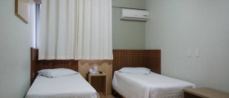 Quarto de hotel com duas camas de solteiro, cabeceira de madeira, mesa de cabeceira com telefone e controle remoto, e aparelho de ar condicionado na parede.