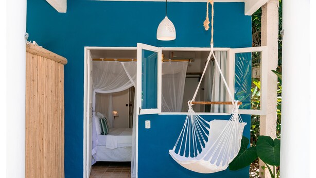 Quarto com parede azul, cama branca com dossel, rede e luminária pendente.