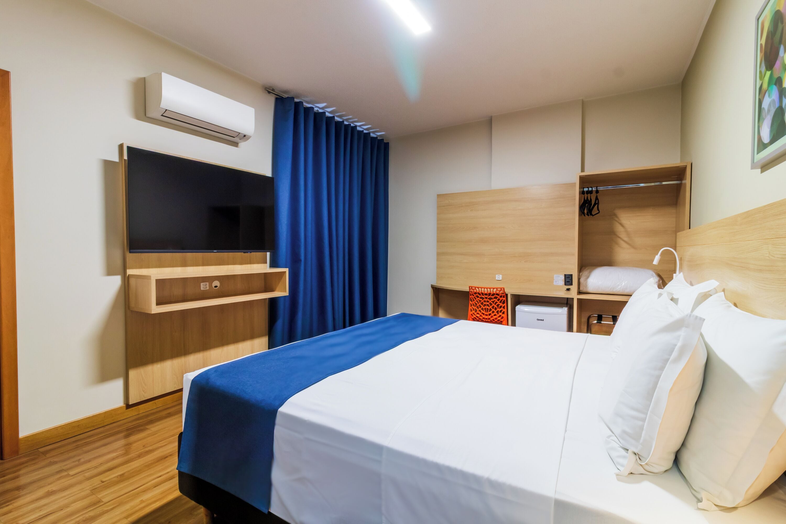 Quarto de hotel com cama, televisão, guarda-roupa, secretária e ar condicionado.