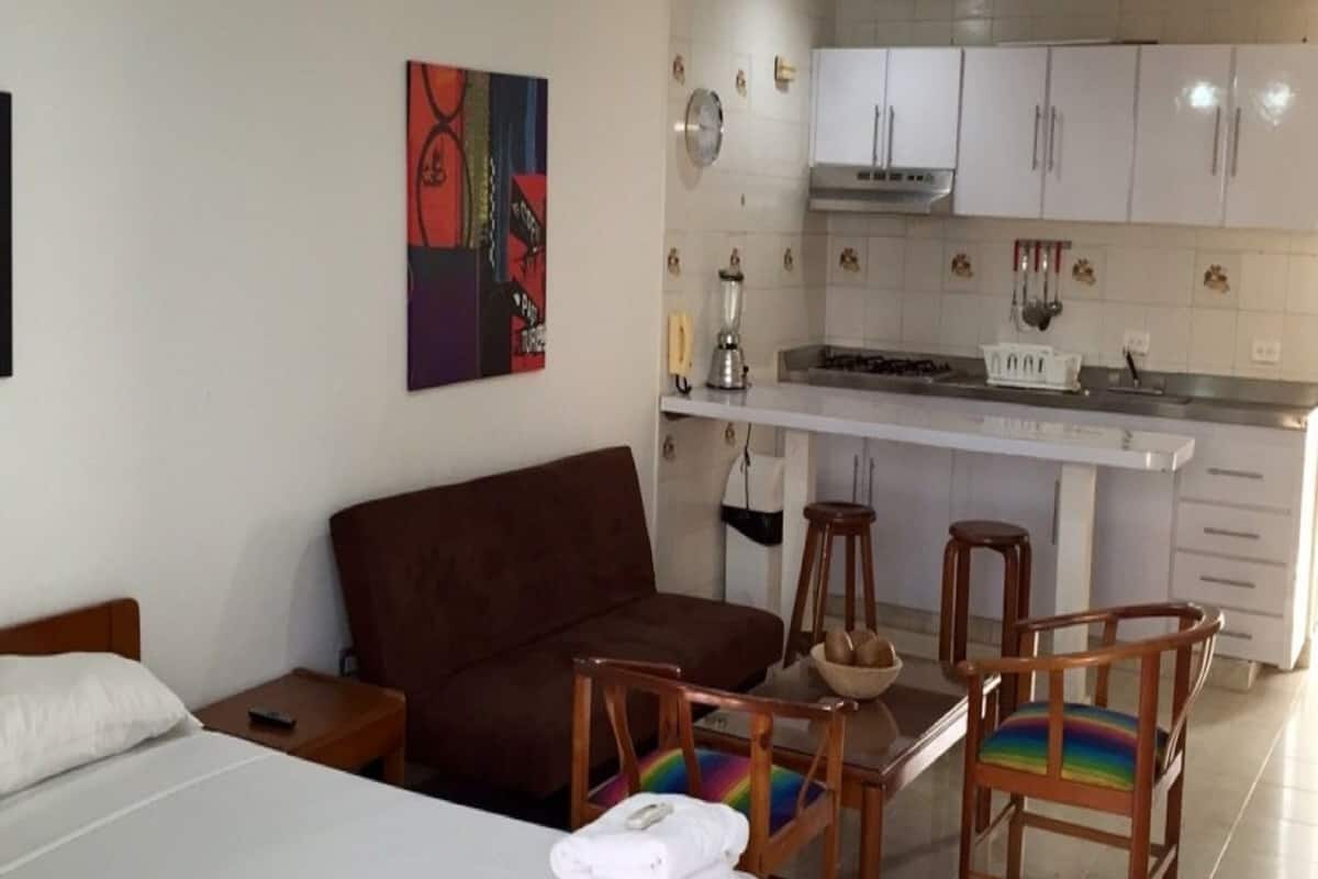 Quarto de hotel com cama, sofá castanho, pequena mesa com cadeiras, área de cozinha com armários brancos e fogão, e uma pintura abstrata colorida na parede.