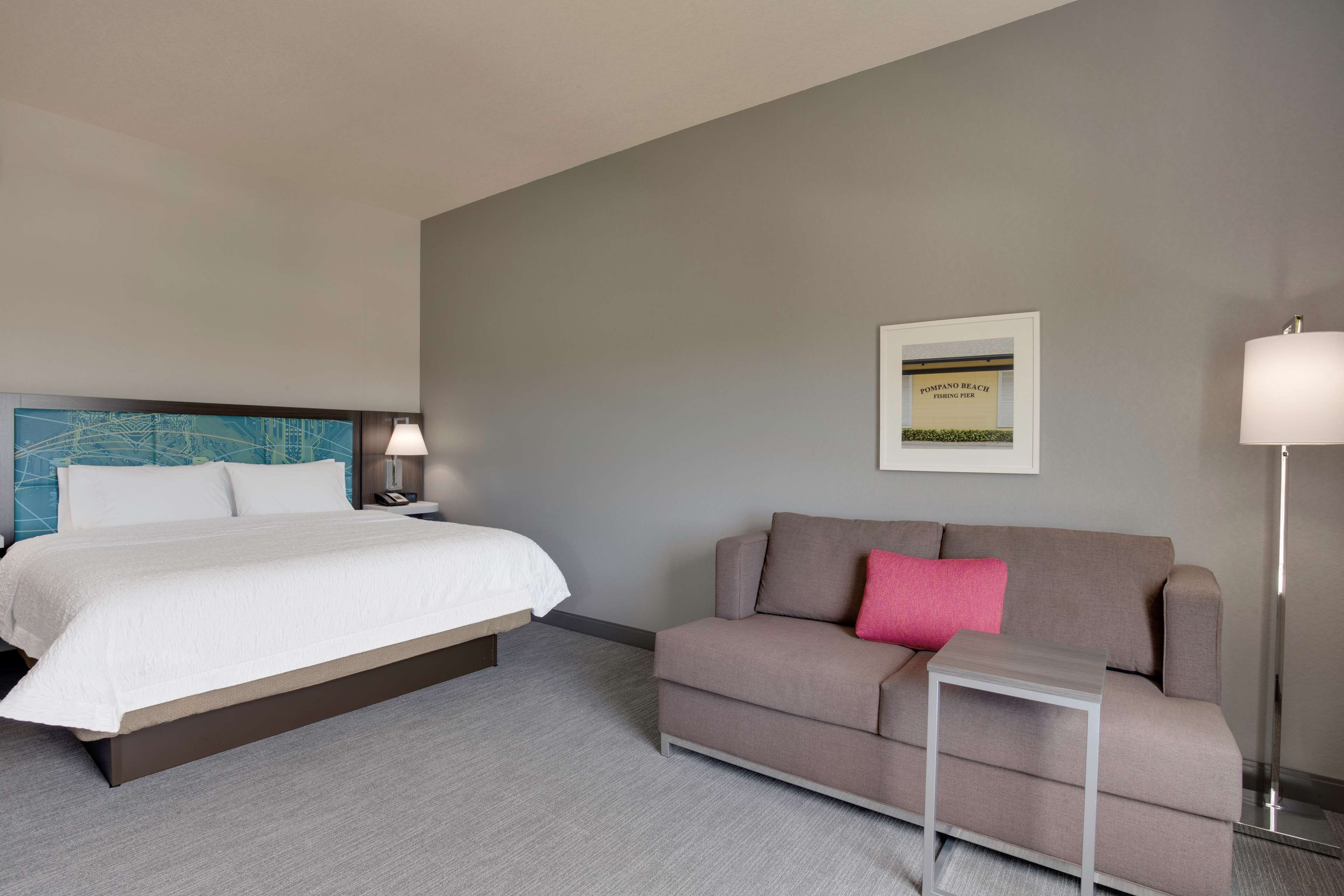 Quarto de hotel com cama, sofá, mesa pequena, abajur e quadro na parede.