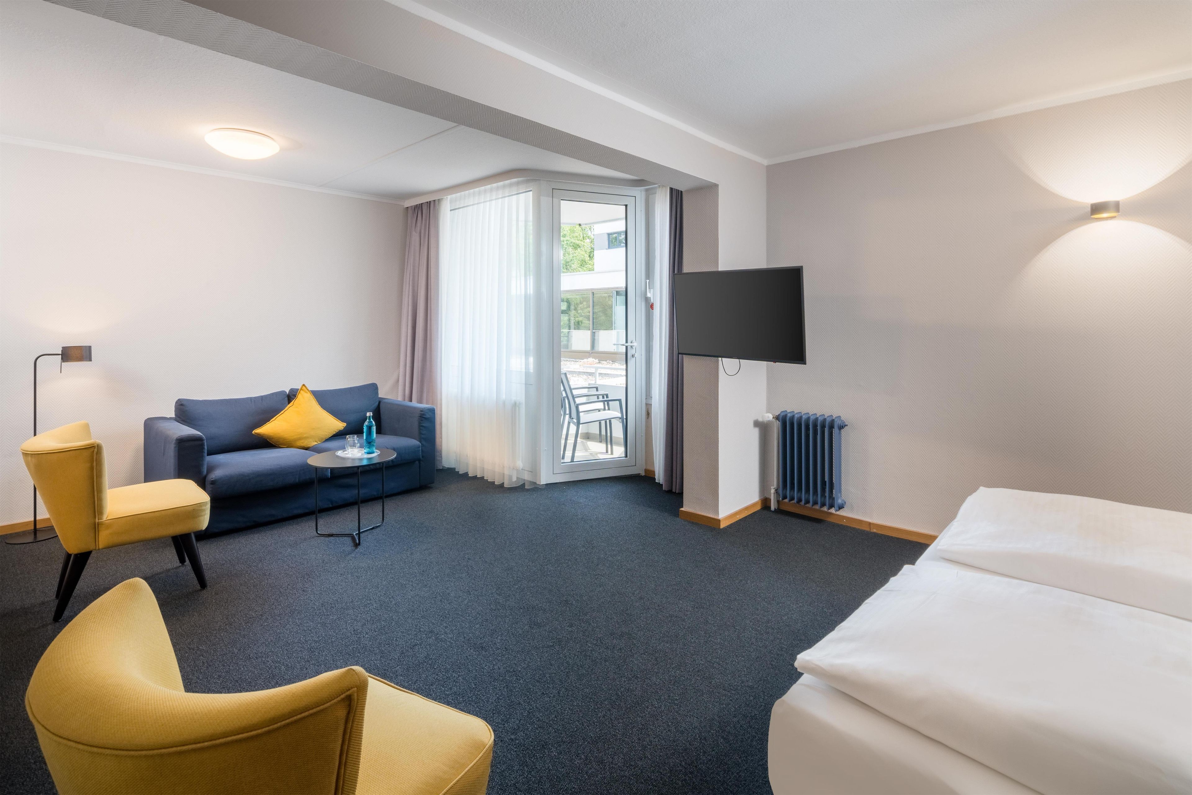 Quarto de hotel com cama, sofá, poltrona, mesinha, televisão e radiador.