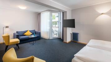 Quarto de hotel com cama, sofá, poltrona, mesinha, televisão e radiador.