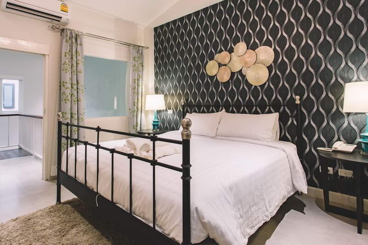 Quarto com estrutura de cama preta em metal, roupa de cama branca, parede estampada e uma pequena mesa de cabeceira com abajur.