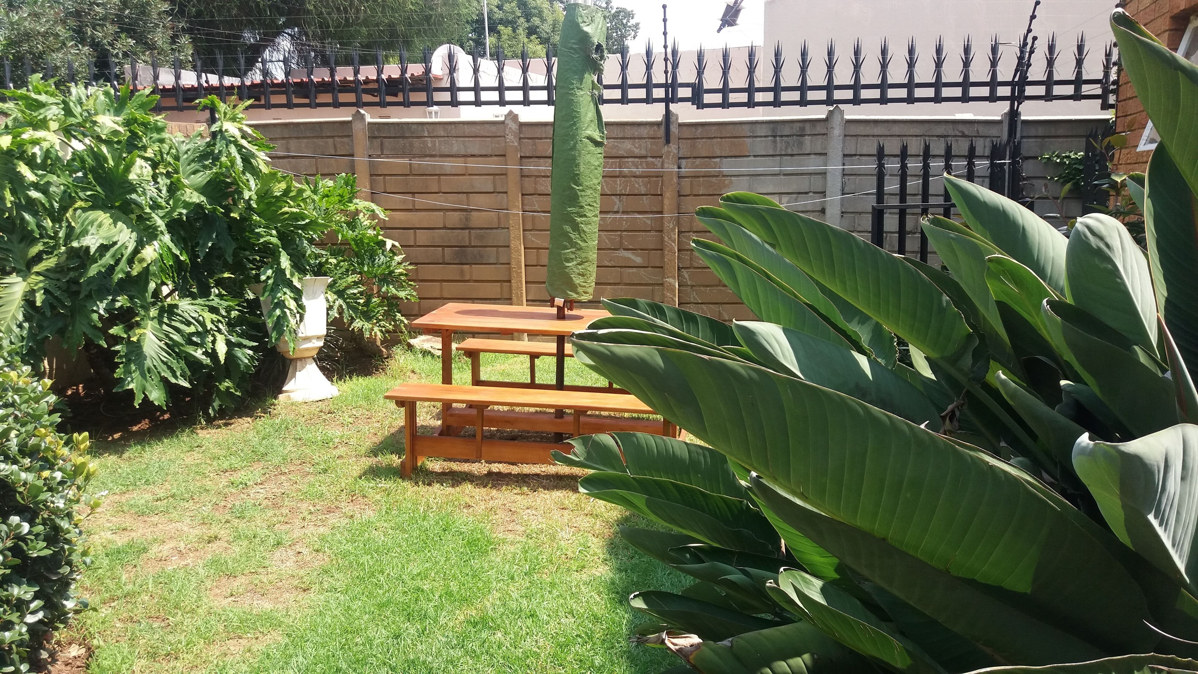Uma área de pátio com mesa e bancos de madeira, rodeada por plantas verdes exuberantes e uma parede de tijolos.