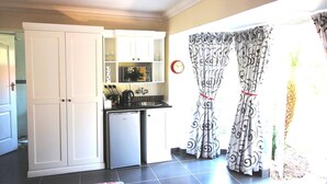 Uma cozinha compacta com armários brancos, uma geladeira pequena e uma pia. Há um relógio na parede e uma cortina estampada sobre a porta.