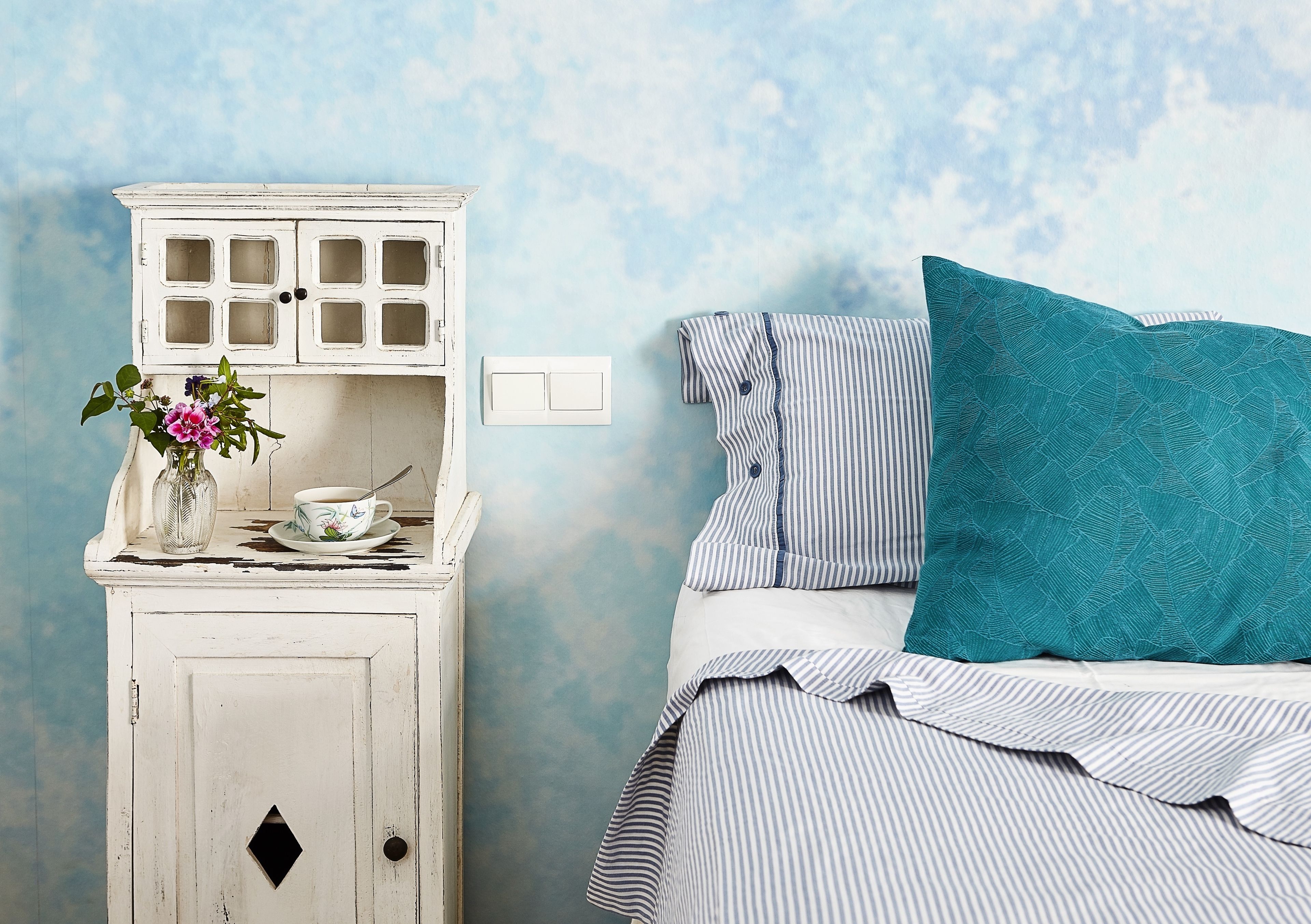 Quarto com cama, mesa de cabeceira e parede com padrão de nuvens.