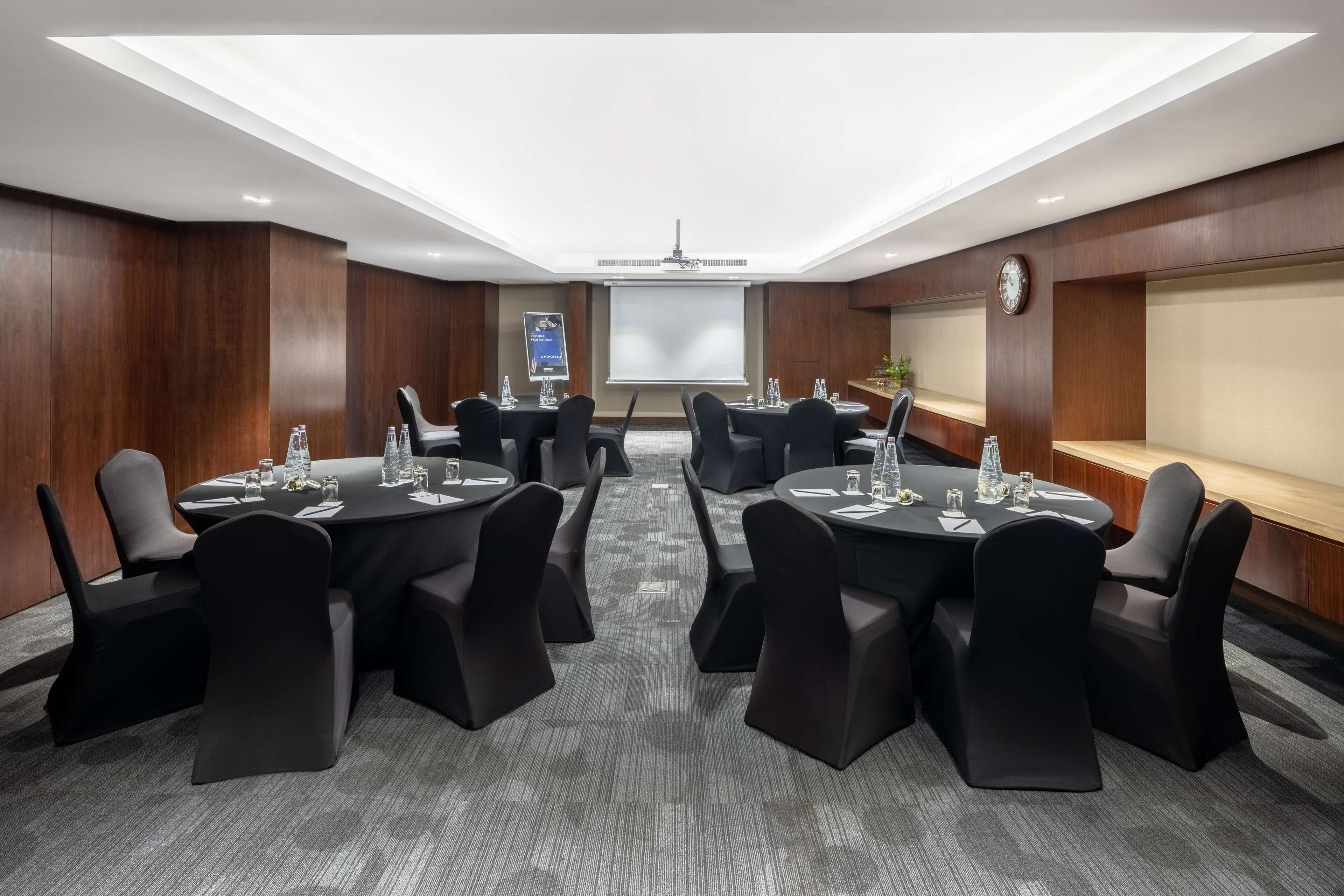 Uma sala de conferências com mesas redondas, cadeiras pretas e uma tela de projetor.