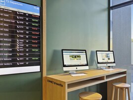 Painel de partidas exibindo voos e números de portão, dois computadores Apple iMac sobre uma mesa de madeira e uma placa com a inscrição 'CONNECT @ MANTRA'.