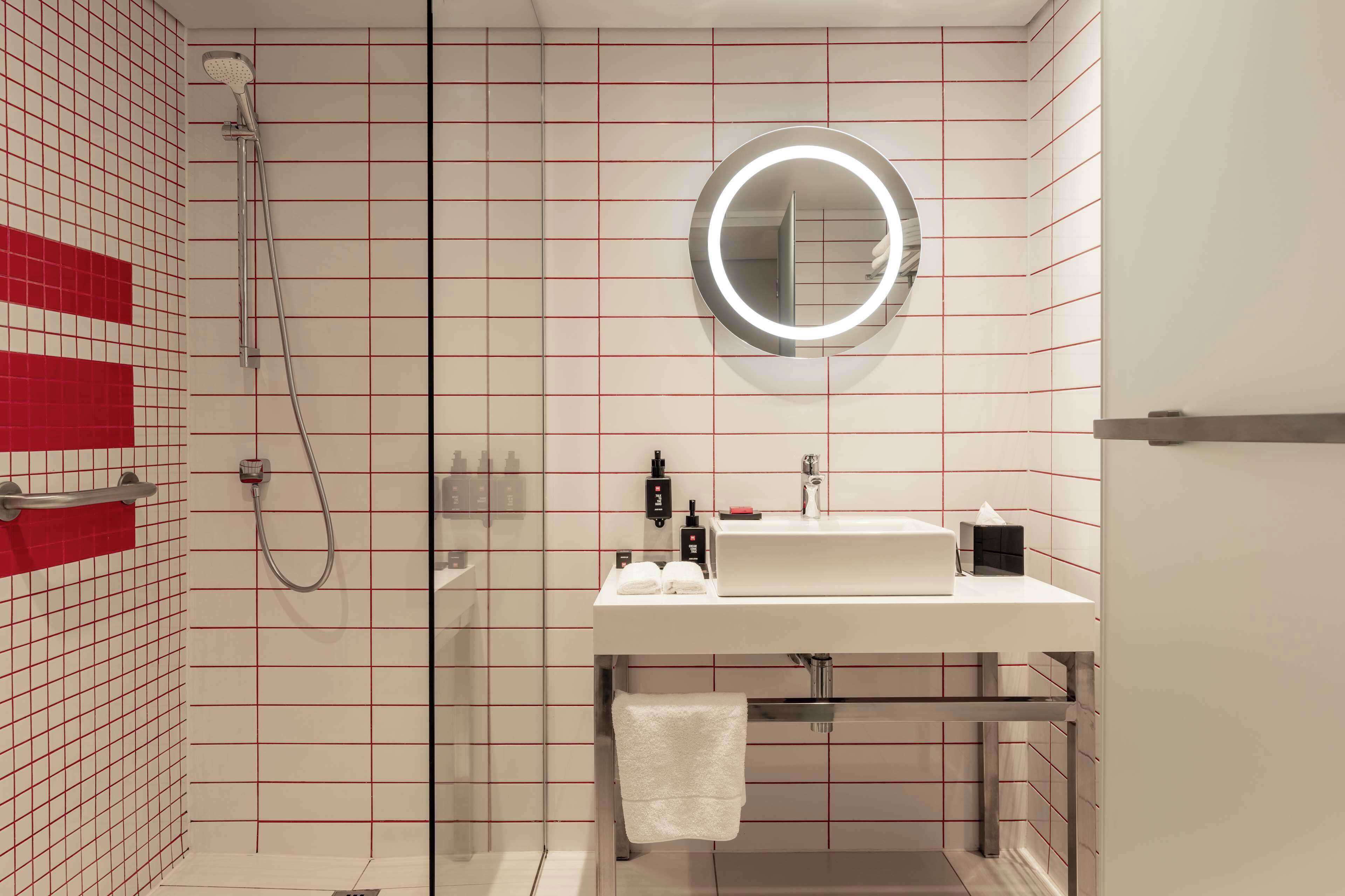 Casa de banho moderna com cabine de duche em vidro, lavatório branco com espelho redondo e paredes com azulejos vermelhos.