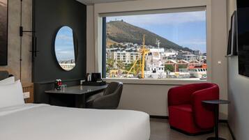 Quarto de hotel com cama, escrivaninha, cadeira, mesa pequena, televisão e grande janela vista para a cidade.