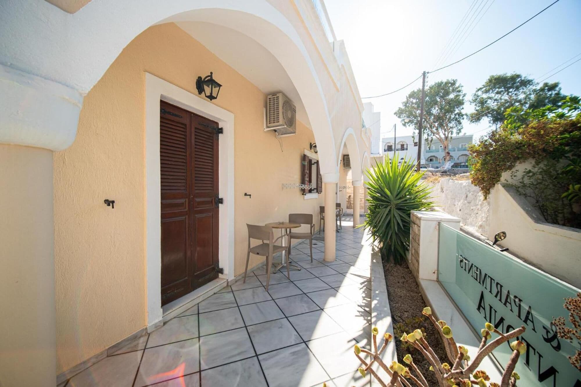 Uma área externa coberta com piso de azulejos, uma porta de madeira e um pequeno espaço de estar com cadeiras e mesas.
