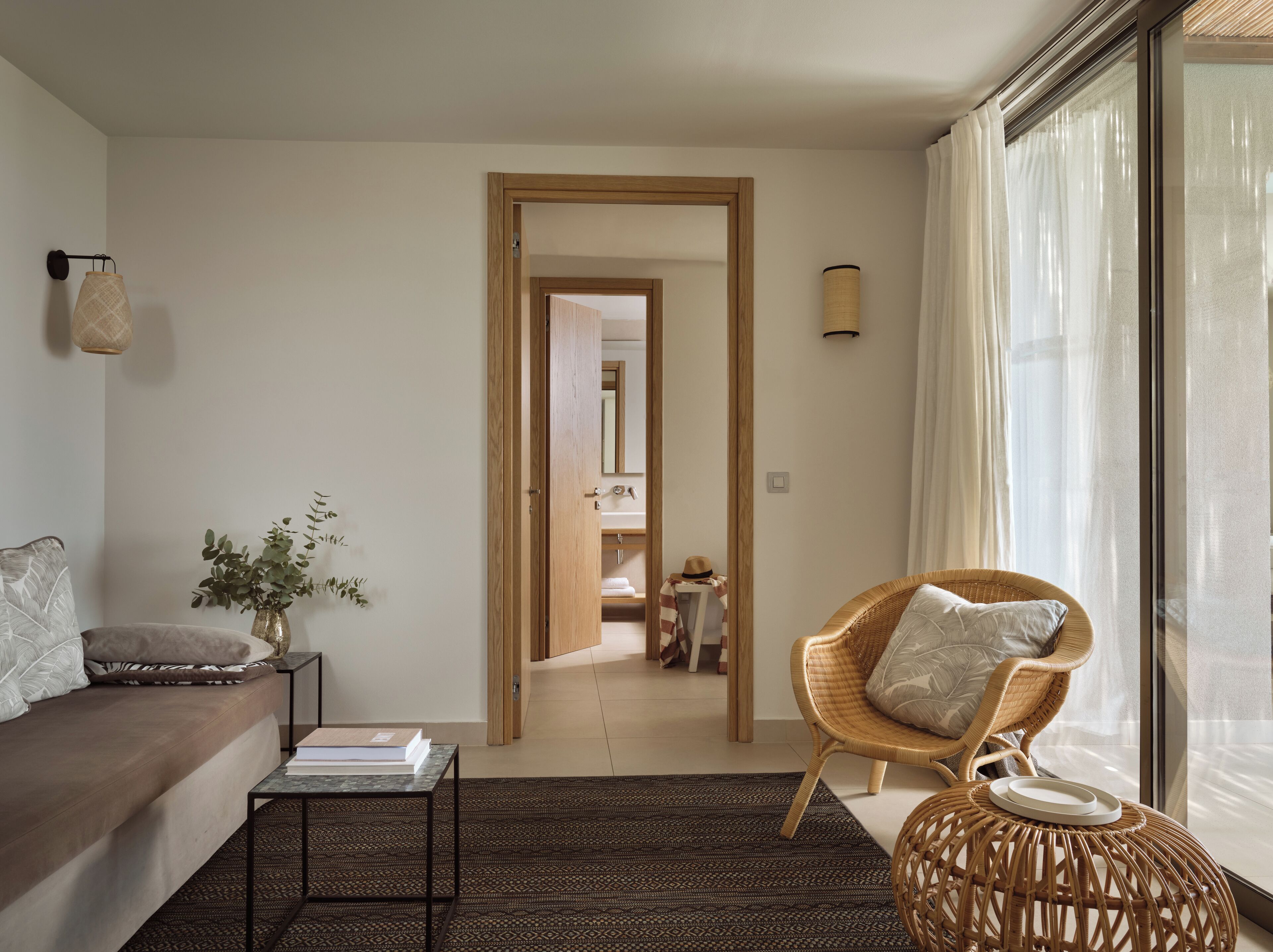 Uma sala de estar moderna com um sofá, uma poltrona de vime, uma mesinha e uma janela grande com cortinas leves.