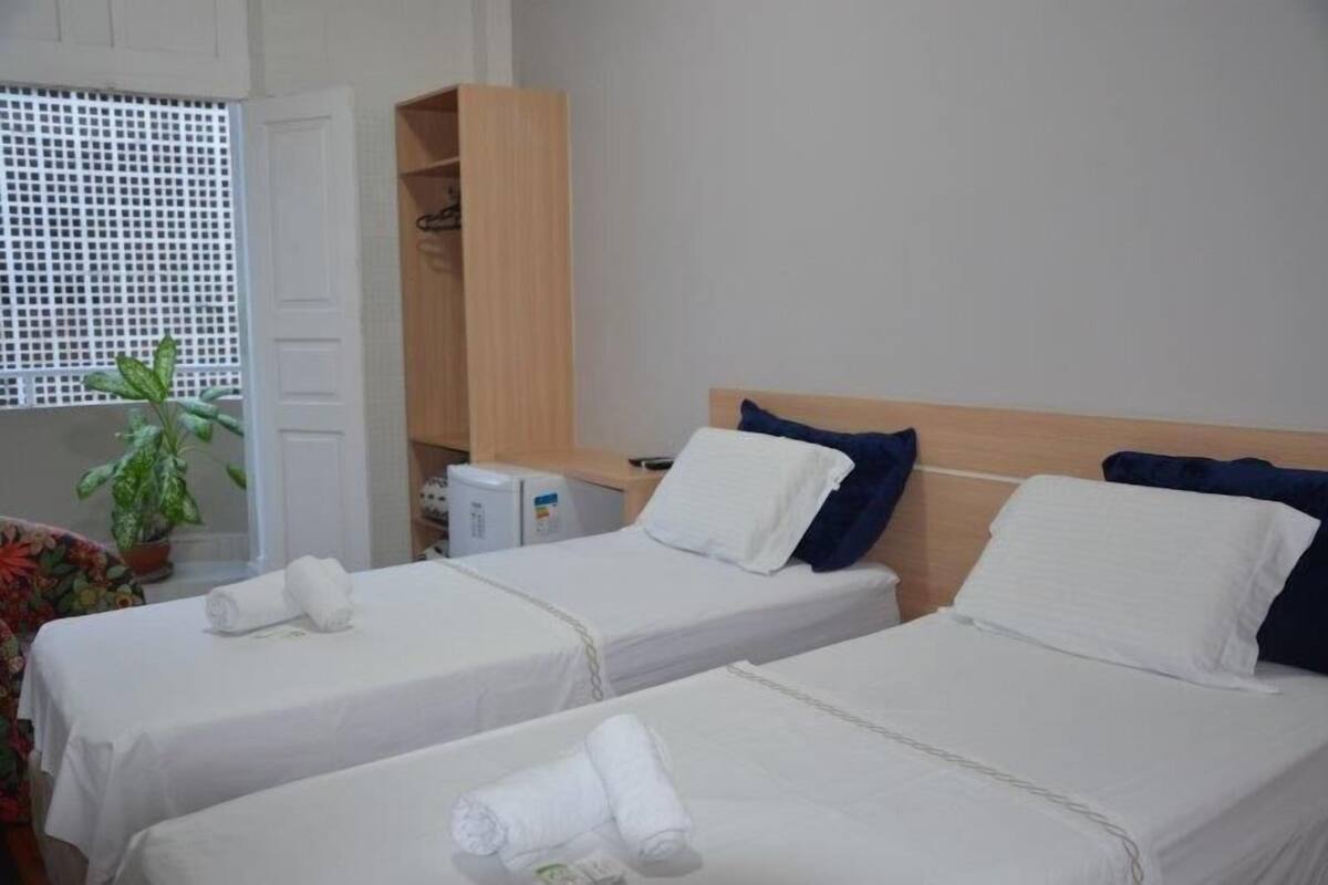 Quarto de hotel com duas camas de solteiro, um guarda-roupa de madeira, uma mesinha com uma planta e uma janela com grade branca.