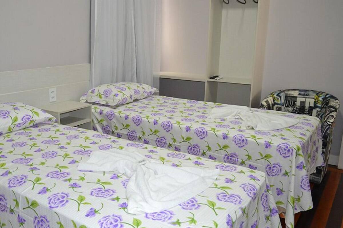 Quarto de hotel com duas camas, cada uma com colcha florida, mesa de cabeceira e uma cadeira.