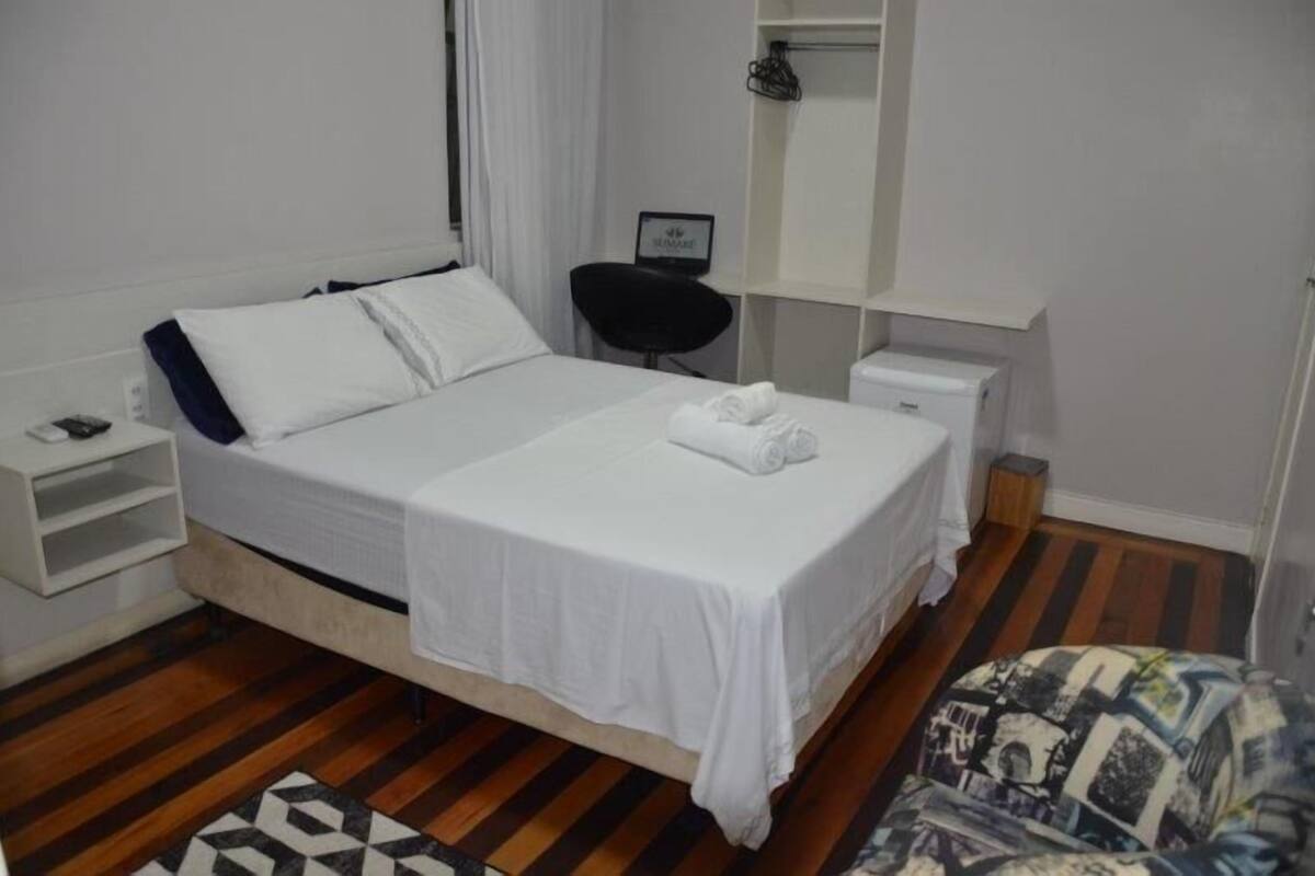 Cama bem arrumada com lençóis brancos, mesa de cabeceira com abajur, cadeira e prateleira pequena.