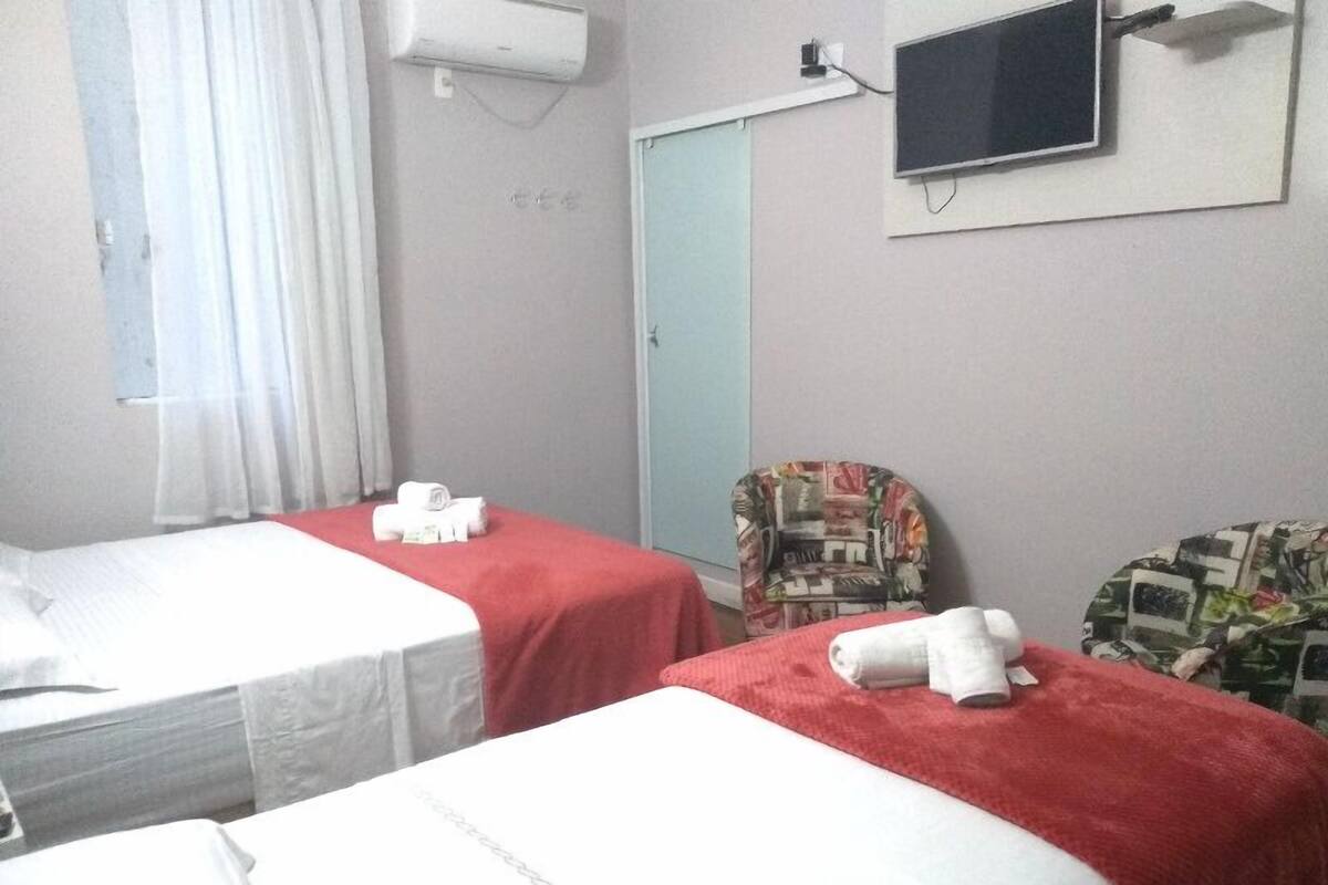 Quarto de hotel com duas camas, cada uma com um cobertor vermelho e almofadas brancas, uma TV fixada na parede e uma poltrona com padrão.