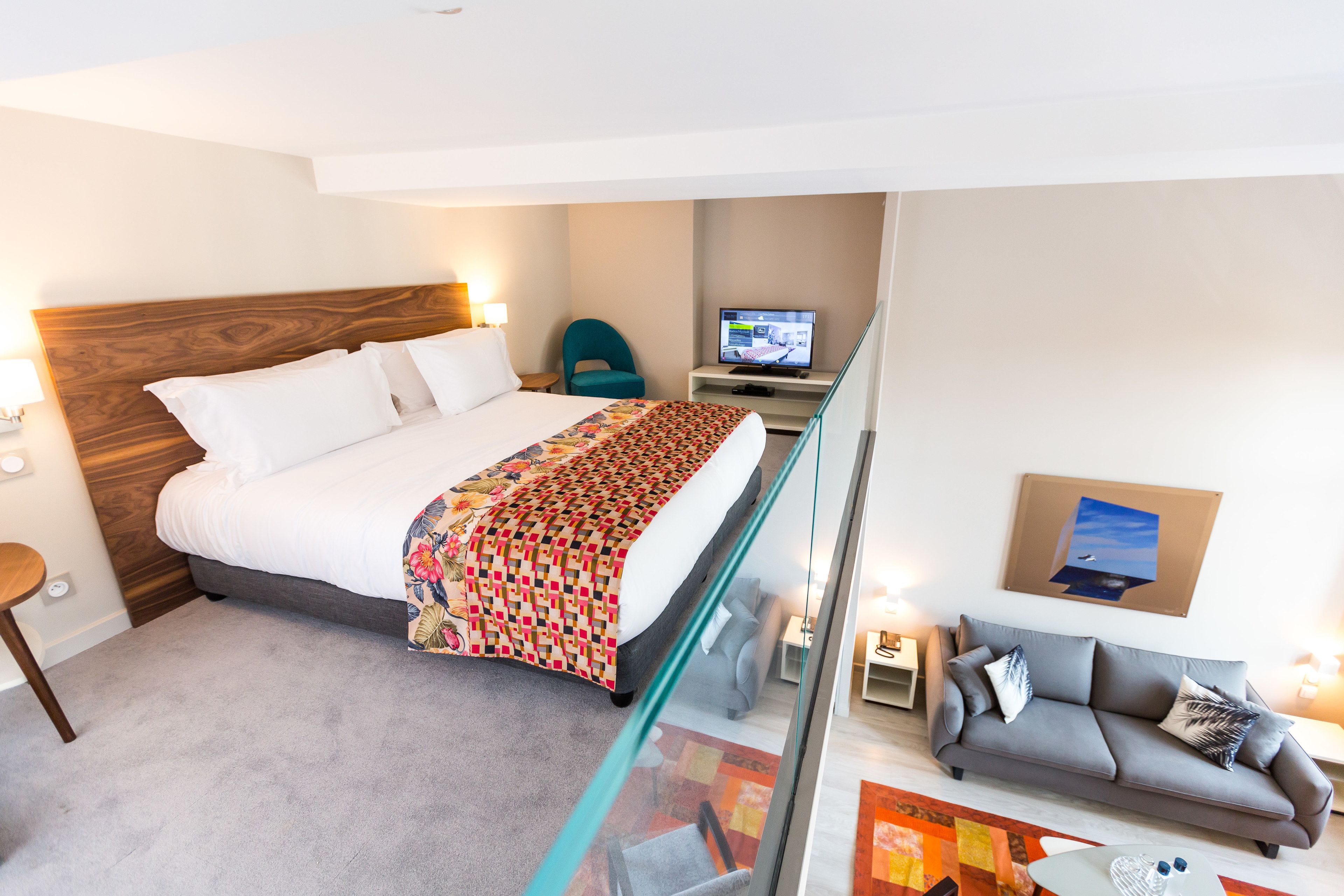 Quarto de hotel com cama, televisão, sofá e guarda-corrimão de vidro.