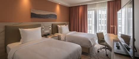 Quarto de hotel com duas camas, uma mesa de trabalho com cadeira, uma luminária de mesa, uma TV de tela plana e um quadro na parede.