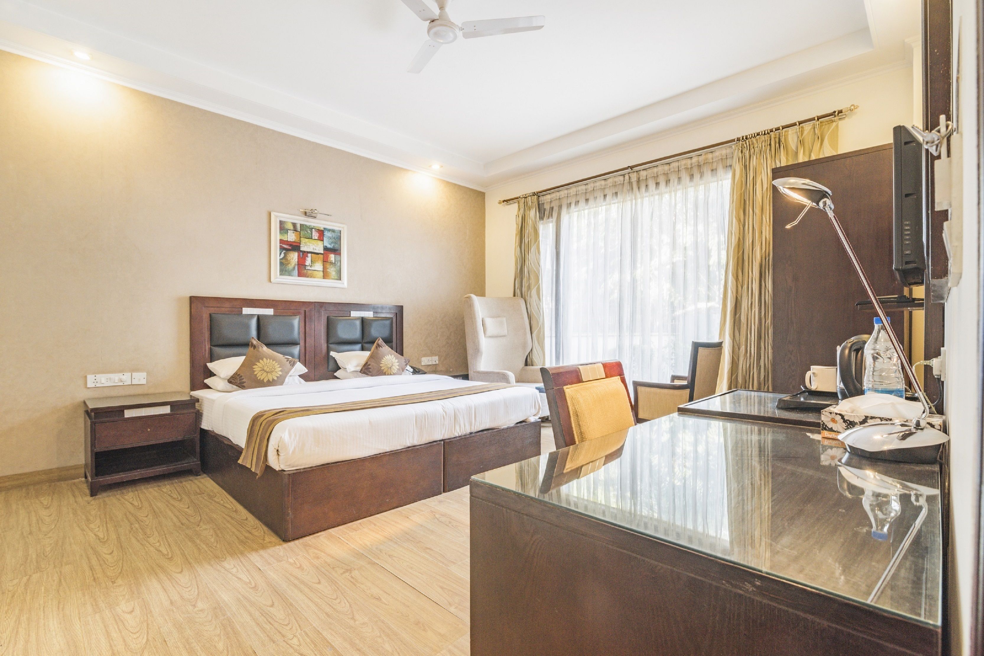 Quarto de hotel com cama, mesa de cabeceira, escrivaninha, cadeira e uma janela grande com cortinas.