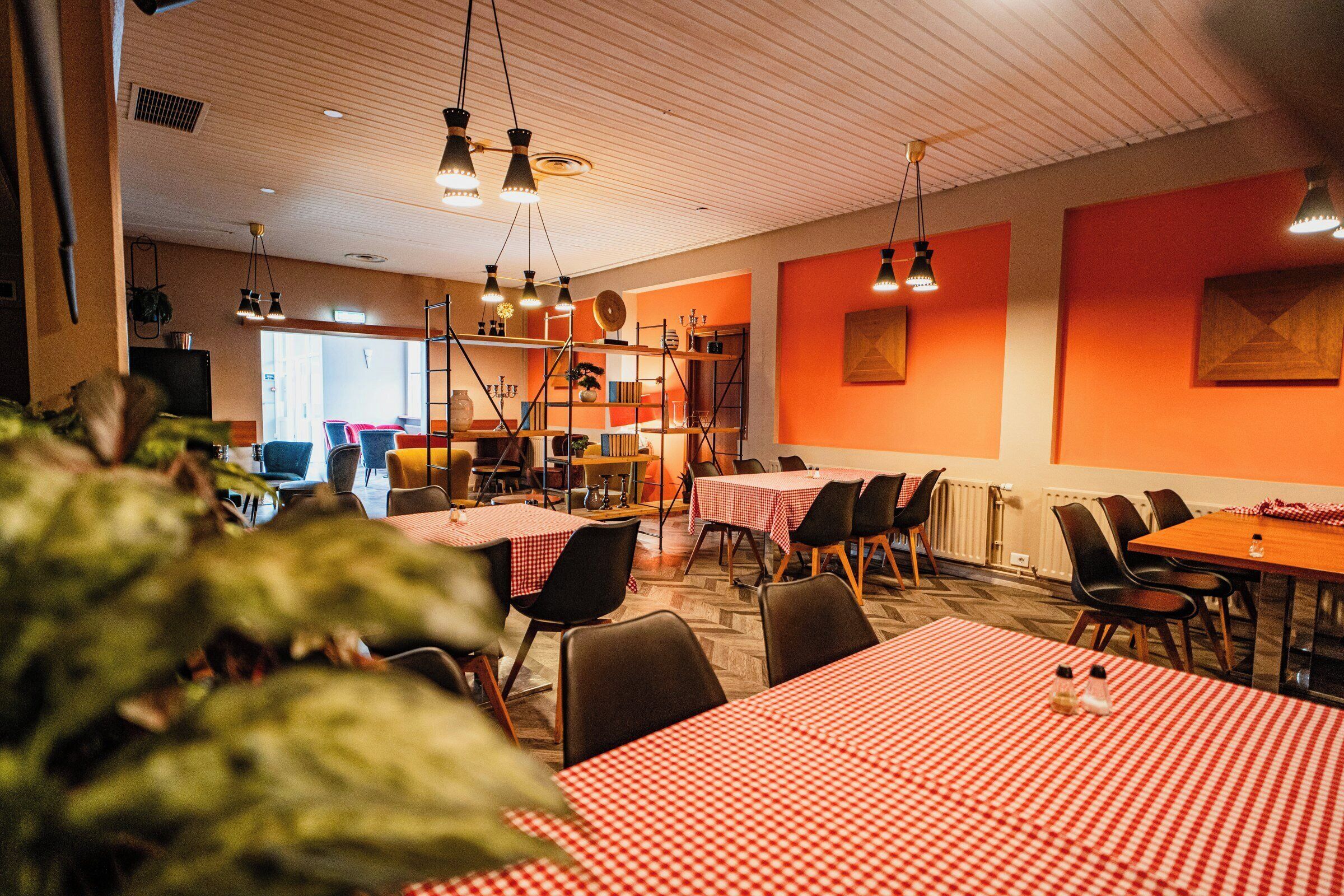 Restaurante com toalhas de mesa xadrez vermelho e branco, cadeiras pretas e luminárias pendentes.