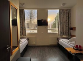 Quarto de hotel com duas camas, uma televisão, uma janela com vista para árvores e uma mesa de madeira com chávenas e um tabuleiro.