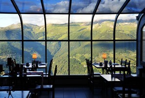 Uma sala de jantar com mesas e cadeiras, grandes janelas com vista para montanhas e vales verdejantes.