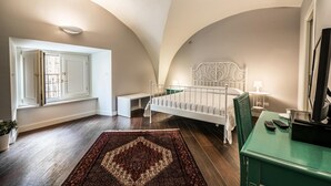Quarto com cama branca, escrivaninha verde, televisão e janela com vista.