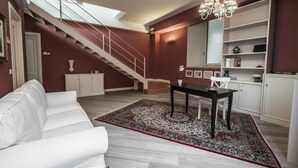 Uma sala de estar moderna com um sofá branco, uma mesa de centro preta e um tapete estampado. Uma escada com corrimão de vidro leva a um andar superior.