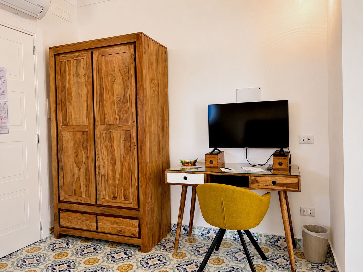 Quarto com guarda-roupa de madeira, escrivaninha com televisão, uma cadeira amarela e ladrilhos no chão com padrão.