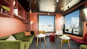 Quarto de hotel moderno com um sofá verde, um banquinho vermelho, uma mesa redonda branca e uma janela grande com vista para uma paisagem nevada.
