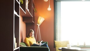 Uma mulher lendo um livro num quarto com estante, candeeiro de chão e janela com cortinas.