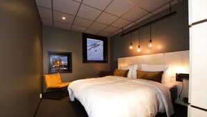 Quarto de hotel moderno com uma cama grande, vista de uma montanha nevada pela janela e um pequeno espaço de estar com uma poltrona.