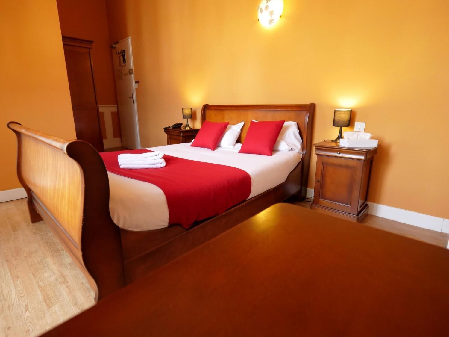 Quarto de hotel com cama de madeira, roupa de cama vermelha e branca, duas mesas de cabeceira com abajures e um criado-mudo de madeira.