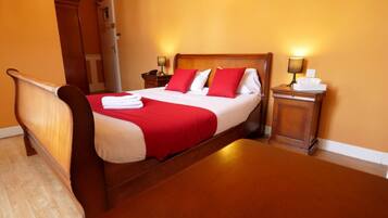 Quarto de hotel com cama de madeira, roupa de cama vermelha e branca, duas mesas de cabeceira com abajures e um criado-mudo de madeira.