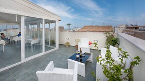 Um terraço no telhado com móveis de vime branco, uma área de refeições com paredes de vidro e plantas em vasos.