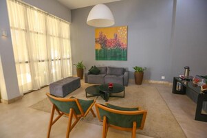 Sala de estar com sofá cinza, mesa de centro e duas poltronas. Há um quadro na parede e uma janela grande com cortinas.