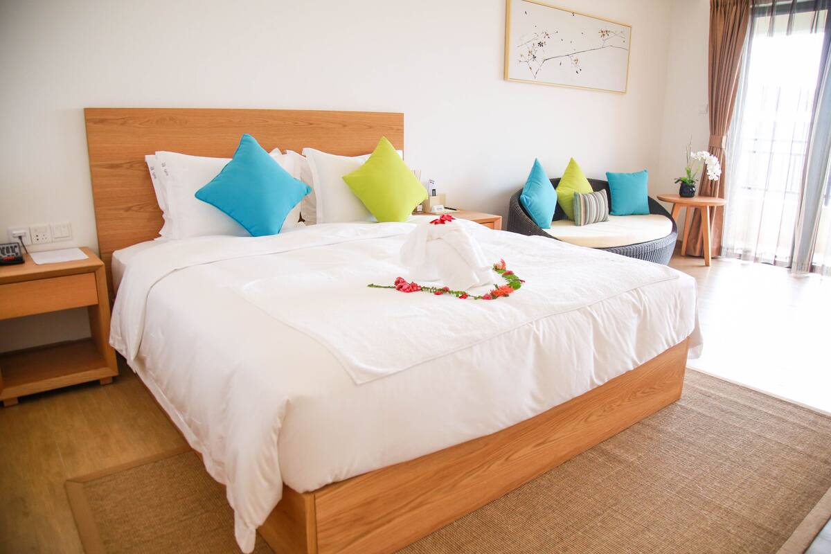 Uma cama bem arrumada com cabeceira de madeira, uma mesinha de cabeceira com abajur e uma pequena mesa com uma planta.