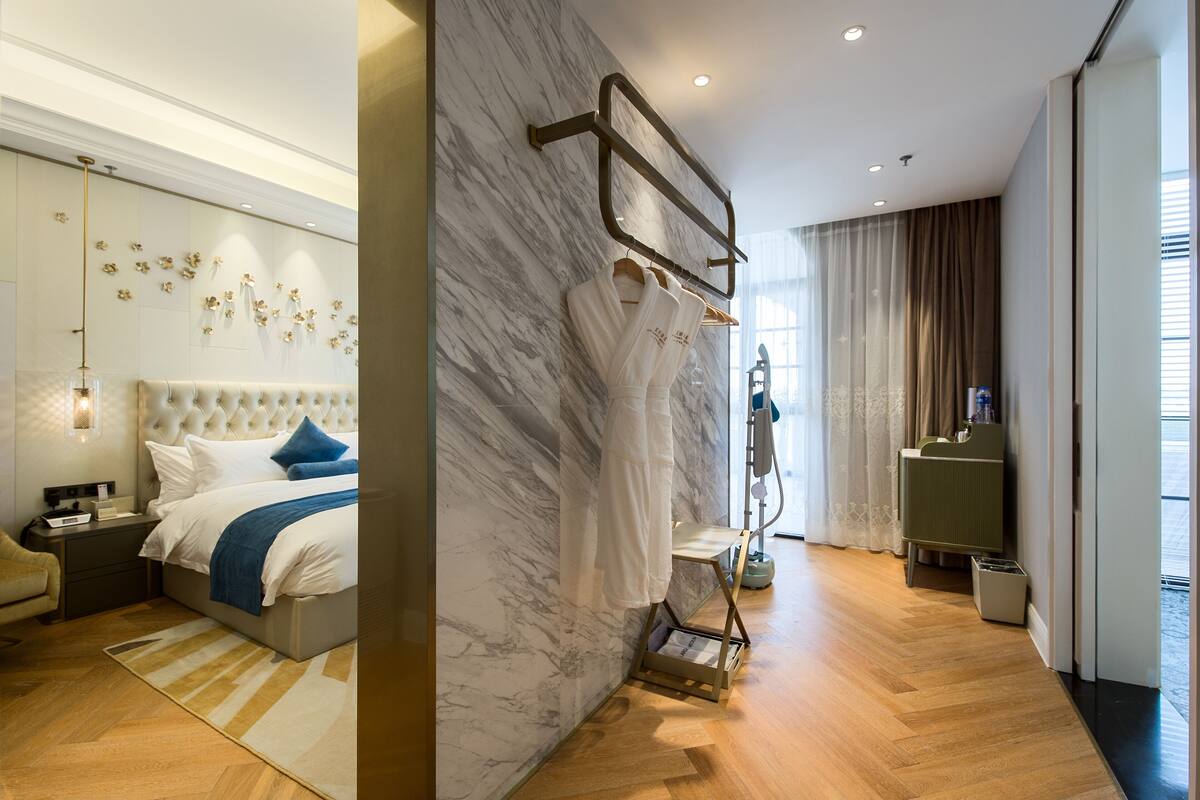 Quarto de hotel moderno com parede de mármore, cama com cabeceira estofada, mesa de cabeceira, cabideiro suspenso e pequena mesa de cabeceira com abajur.