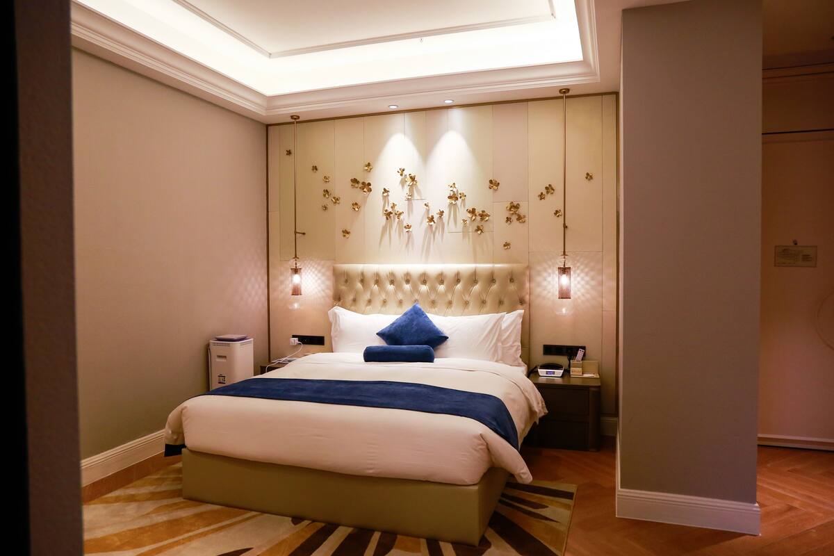 Quarto de hotel com uma cama grande, cabeceira estofada, mesa de cabeceira e luminária fixada na parede.