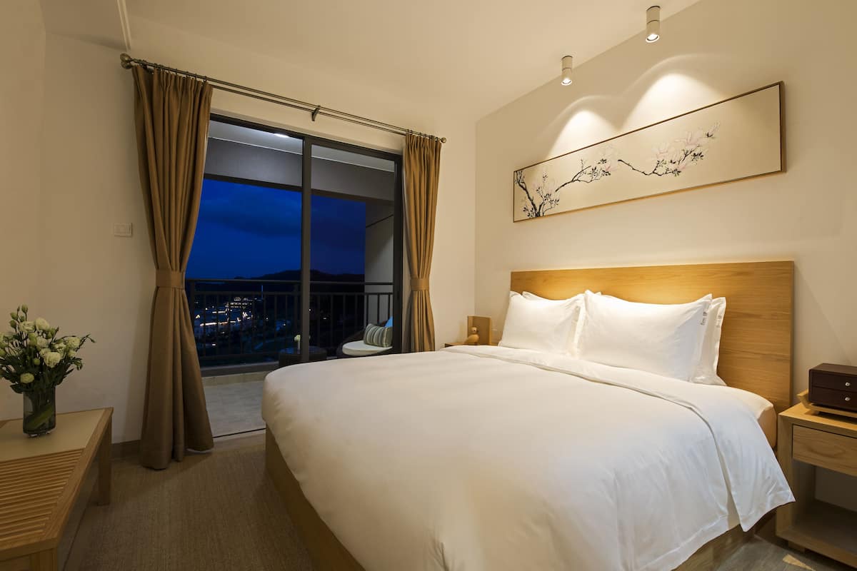 Quarto de hotel com uma cama grande, uma mesa de cabeceira, uma mesinha de apoio com um vaso de flores, vista da cidade à noite através de uma porta de correr e uma obra de arte emoldurada na parede.