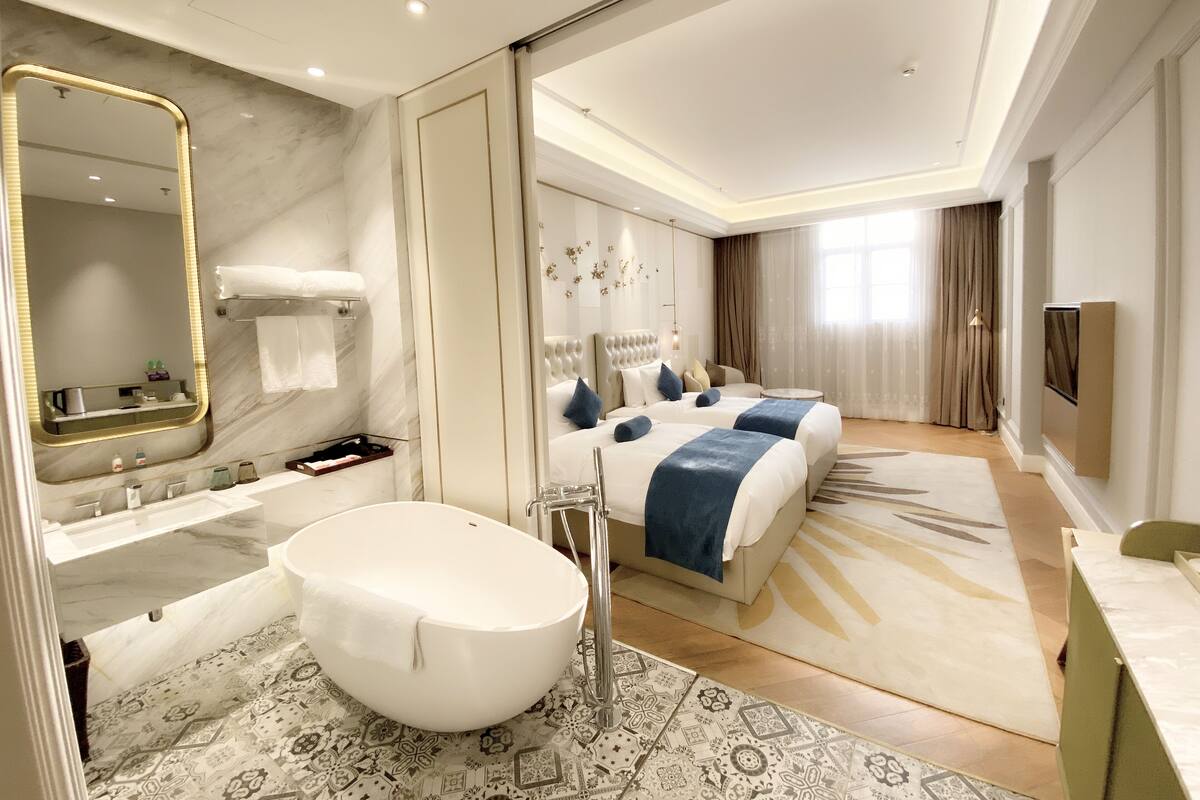 Quarto de hotel moderno com uma cama grande, um espaço de estar separado, uma banheira e um espelho amplo.
