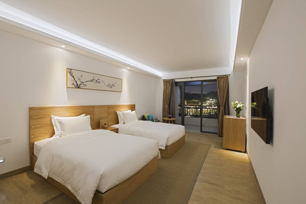 Quarto de hotel com duas camas, varanda com cortinas, televisão fixada na parede e um quadro na parede.