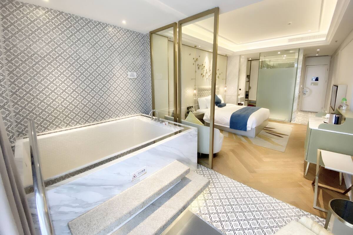 Quarto de hotel moderno com uma banheira grande, uma cama com manta azul, uma escrivaninha e um espelho.