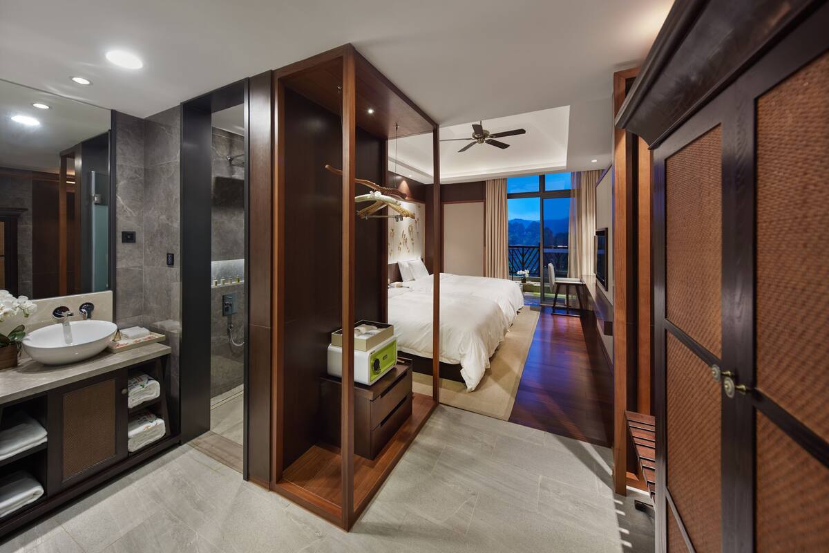 Quarto de hotel moderno com uma cama grande, banheiro com toucador e pia, e varanda com vista.